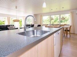 Uma cozinha moderna com uma ilha grande, pia de aço inoxidável e armários de madeira. Há uma sala de jantar com mesa e cadeiras, e uma sala de estar com sofá e cortinas.