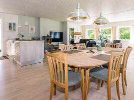 Uma cozinha moderna com ilha central, área de jantar com mesa redonda e cadeiras, e sala de estar com sofá e televisão.