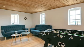 Quarto com mesa de futebol, dois sofás, uma mesa redonda pequena e duas janelas.