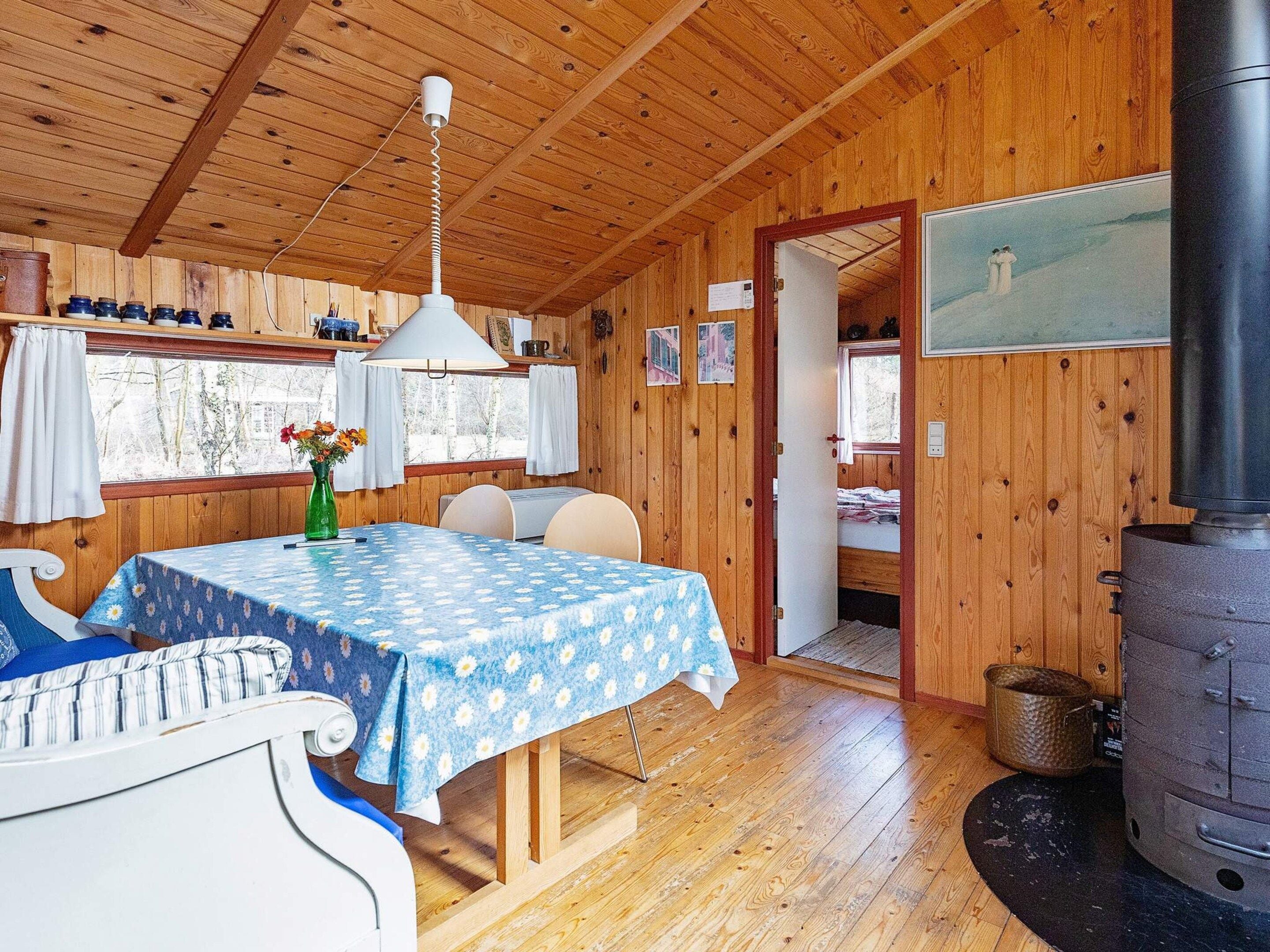Interior de uma cabana de madeira com mesa de jantar, cadeiras, luminária pendente, fogão e janela com cortinas.