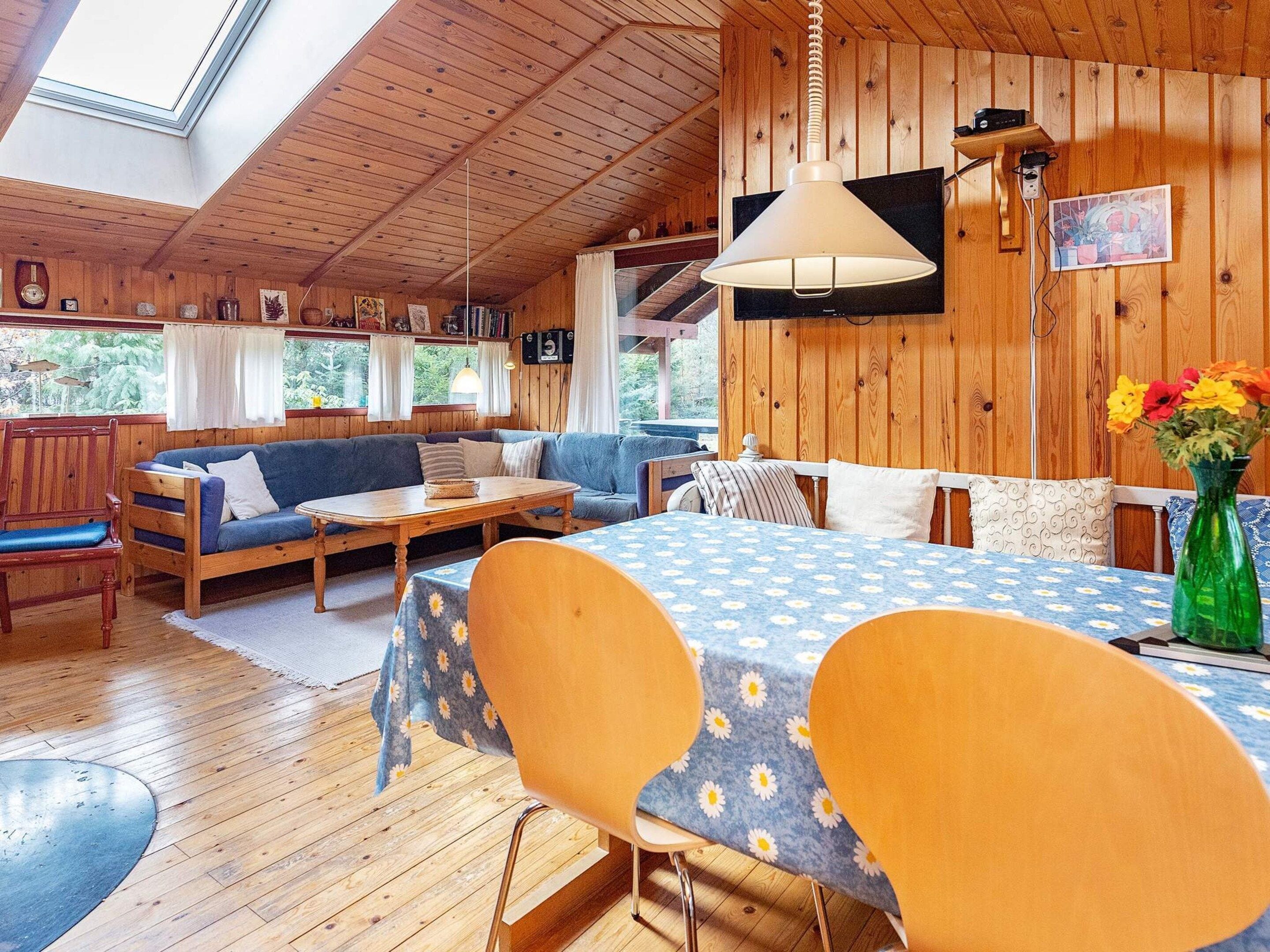 Interior acolhedor de uma cabana com teto de madeira, um sofá, uma mesa de jantar e uma TV fixada na parede.