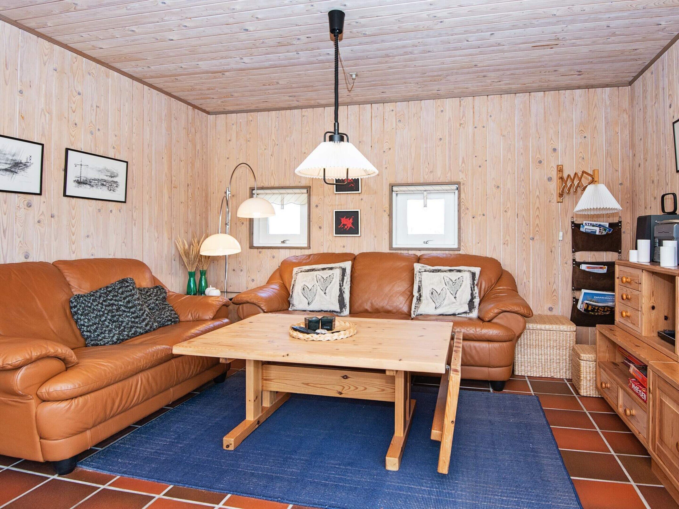 Um quarto aconchegante com um sofá de couro marrom, uma mesa de centro de madeira e um tapete azul. Há duas luminárias fixadas na parede, uma janela com vista e uma prateleira com diversos objetos.