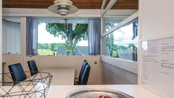 Quarto com mesa, cadeiras, ventilador de teto e janela com vista para árvores e um edifício.
