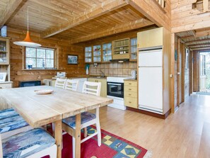 Interior de uma cabana de madeira com mesa de jantar, cadeiras, cozinha com armários, fogão e geladeira.