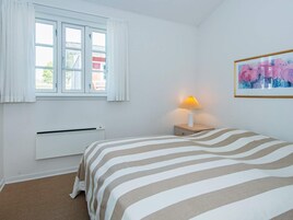 Quarto de hotel com uma cama, radiador, janela com cortinas, mesa de cabeceira com luminária e um quadro na parede.