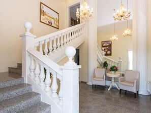 Uma imponente escadaria com balaústres brancos e piso de mármore, que leva a um patamar com duas poltronas e uma pequena mesa.