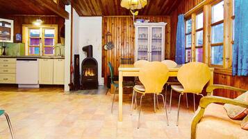 Interior acolhedor de uma cabana com mesa de jantar de madeira, cadeiras, lareira e área de cozinha com armários e pia.