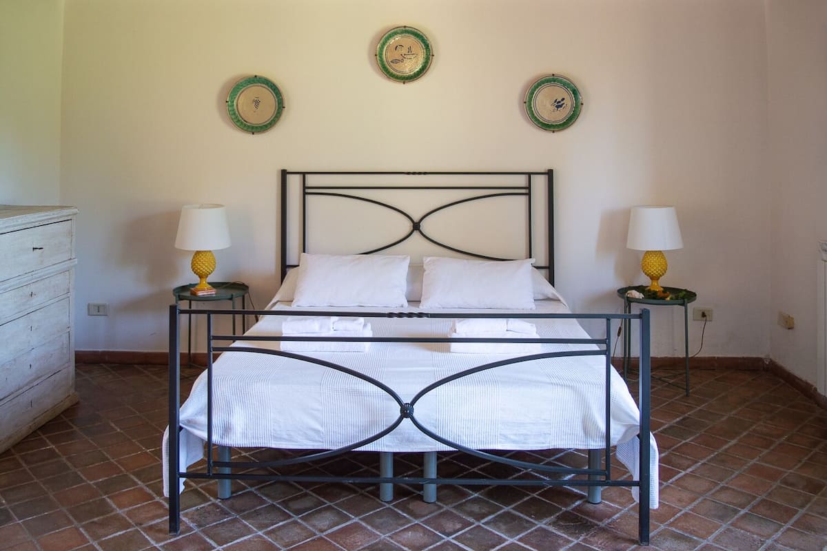 Uma cama com estrutura de metal, roupa de cama branca, duas mesas de cabeceira com abajures e três pratos decorativos na parede.
