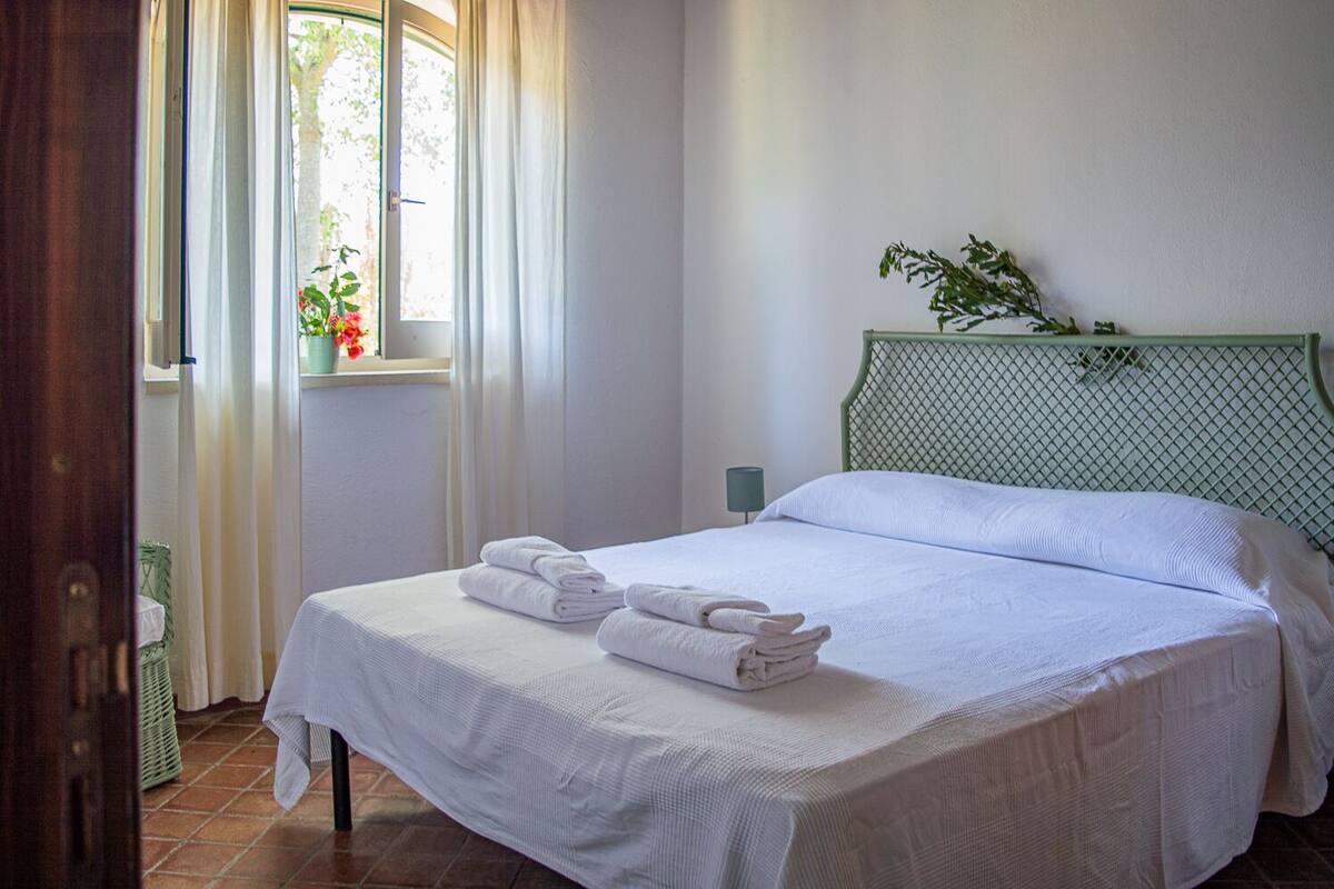 Uma cama bem arrumada com toalhas dobradas, uma janela com cortinas e uma cabeceira verde.