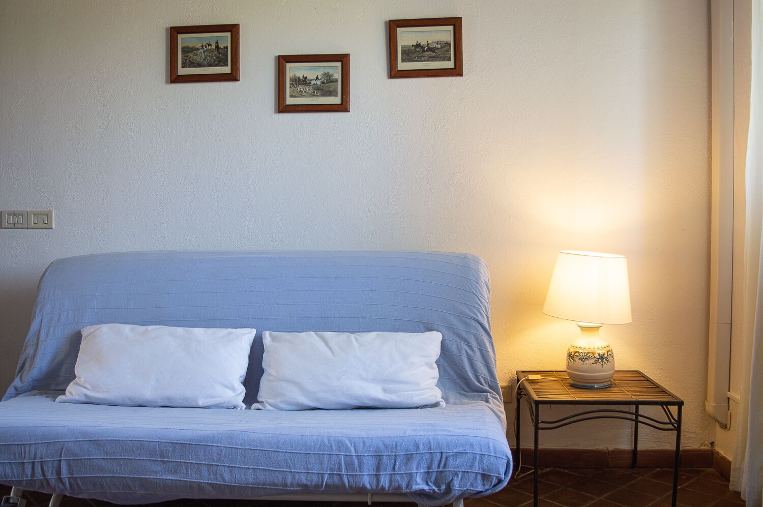 Cama com manta azul e duas almofadas brancas, mesa de cabeceira com abajur e três quadros na parede.