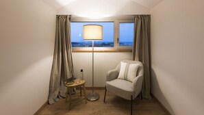 Quarto com vista para o mar, uma lâmpada, uma cadeira e uma pequena mesa com um livro e uma garrafa.