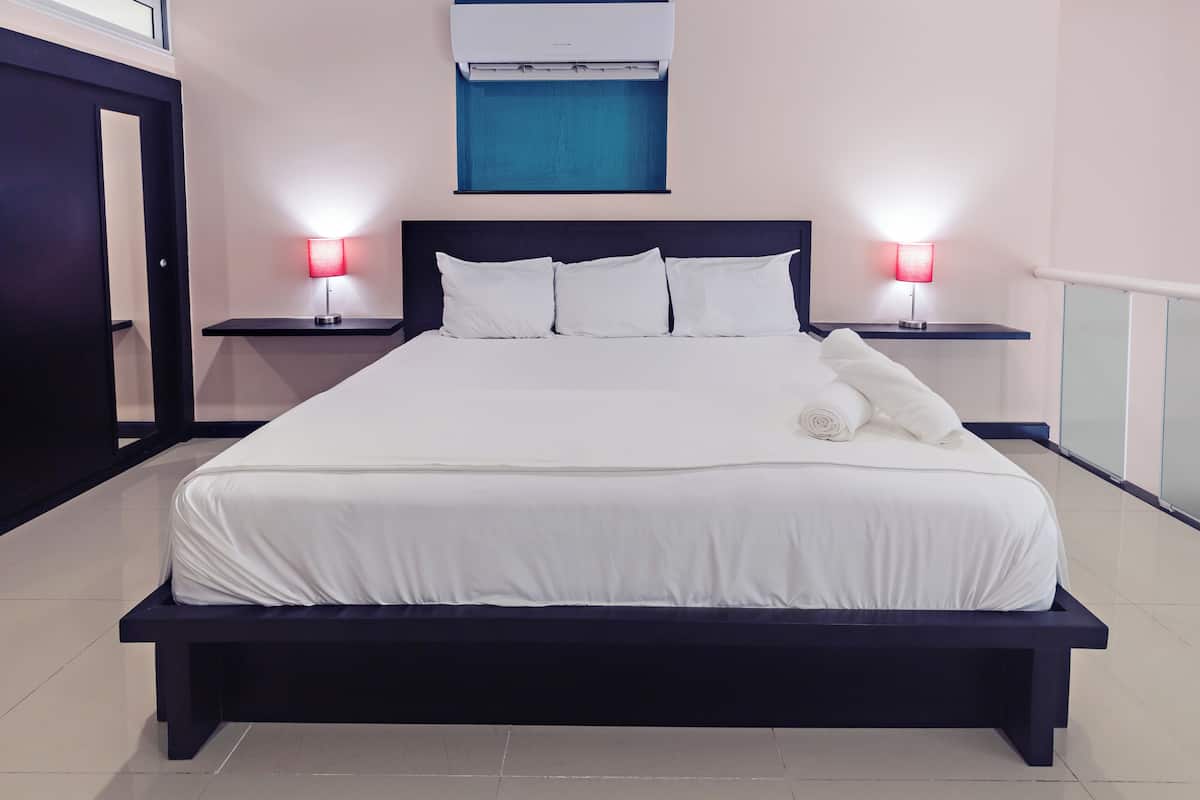 Cama bem arrumada com lençóis brancos, duas luminárias de cabeceira, ar condicionado fixado na parede e janela com cortinas azuis.
