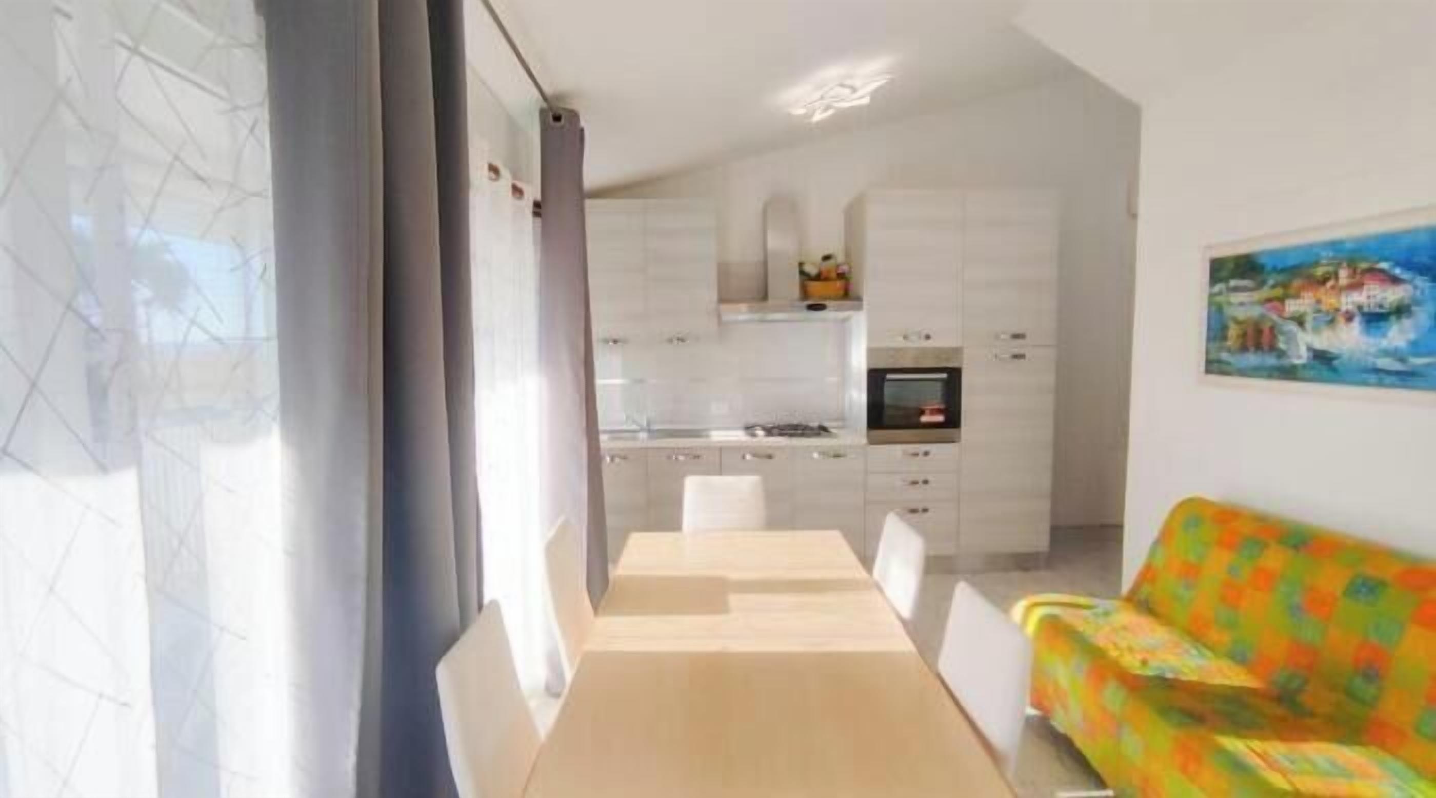Cozinha moderna com armários brancos, fogão e pia. Há uma área de jantar com mesa e cadeiras de madeira, e um sofá colorido.