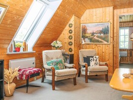 Um quarto aconchegante com paredes de madeira, teto inclinado, uma mesa de jantar, duas poltronas, uma estante de livros e um quadro.