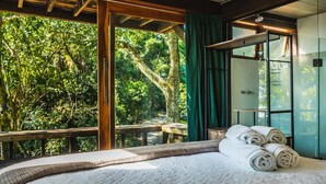Quarto com uma grande janela com vista para uma floresta, cama com toalhas dobradas e um deck de madeira lá fora.