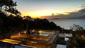 Área de piscina no terraço com deck de madeira e assentos, com vista para um corpo d'água ao pôr do sol.