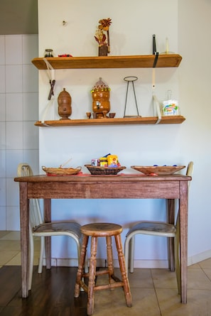 Uma mesa de jantar de madeira com duas cadeiras, uma prateleira acima com utensílios de cozinha e um cesto com alimentos.