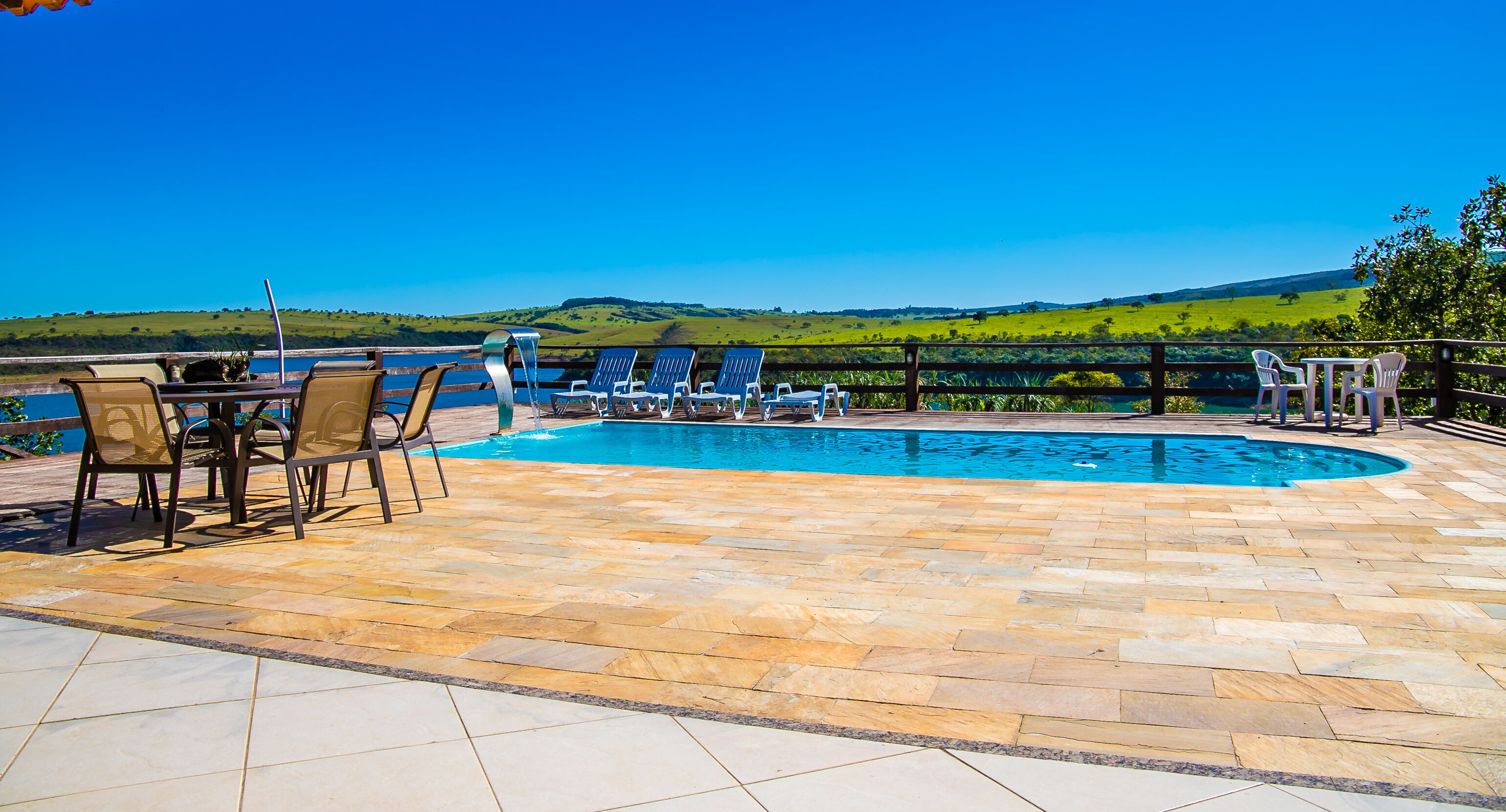 Área da piscina com espreguiçadeiras e uma mesa, rodeada por ladrilhos de pedra, com vista para uma paisagem deslumbrante.