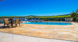 Área da piscina com espreguiçadeiras e uma mesa, rodeada por ladrilhos de pedra, com vista para uma paisagem deslumbrante.