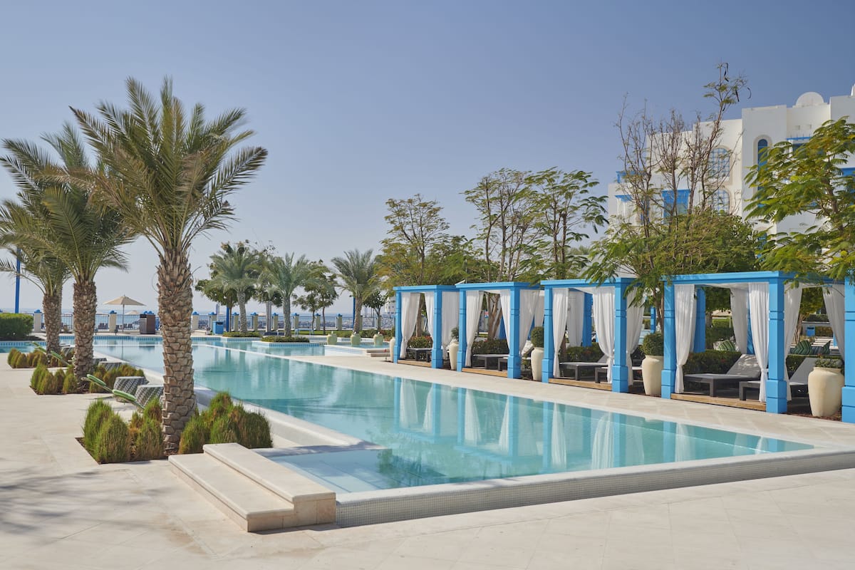 Uma área de piscina com palmeiras, assentos com toldos azul e branco, e vista desobstruída para o mar.
