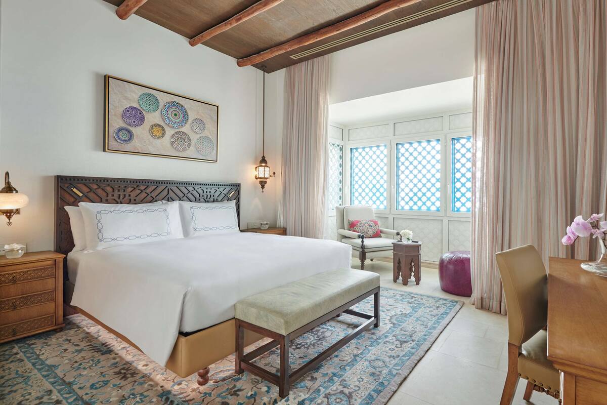 Quarto com uma cama grande, cabeceira de madeira, mesa de cabeceira com abajur, um banco, uma cadeira, uma pequena mesa com um vaso de flores e uma janela com cortinas.