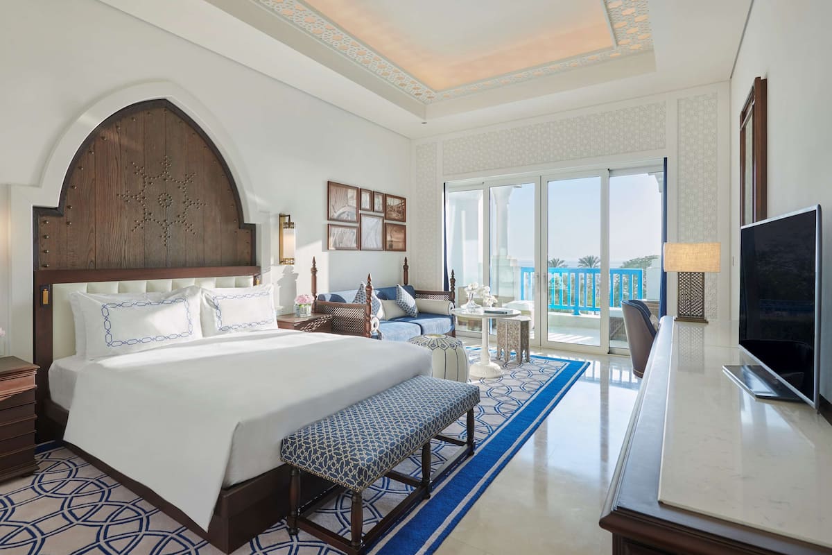 Quarto de hotel com uma cama grande, um banco, uma televisão, varanda com vista e teto decorativo.