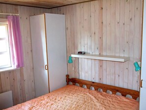 Quarto com guarda-roupa de madeira, cama com colcha laranja, prateleira com relógio e janela com cortinas cor-de-rosa.
