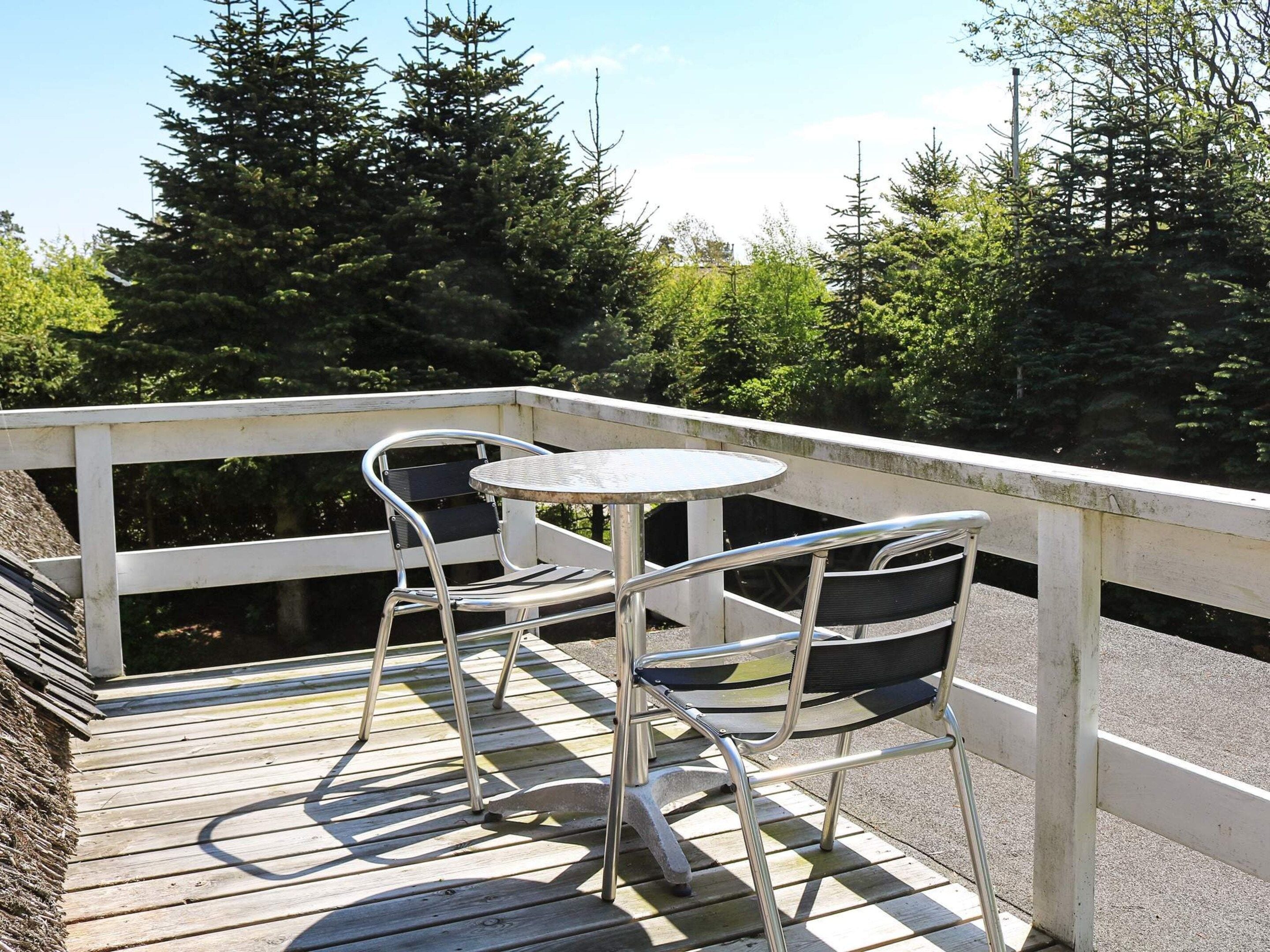Um deck com mesa redonda e duas cadeiras, cercado por guarda-corpo branco, com vista para uma área arborizada.