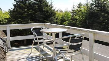 Um deck com mesa redonda e duas cadeiras, cercado por guarda-corpo branco, com vista para uma área arborizada.