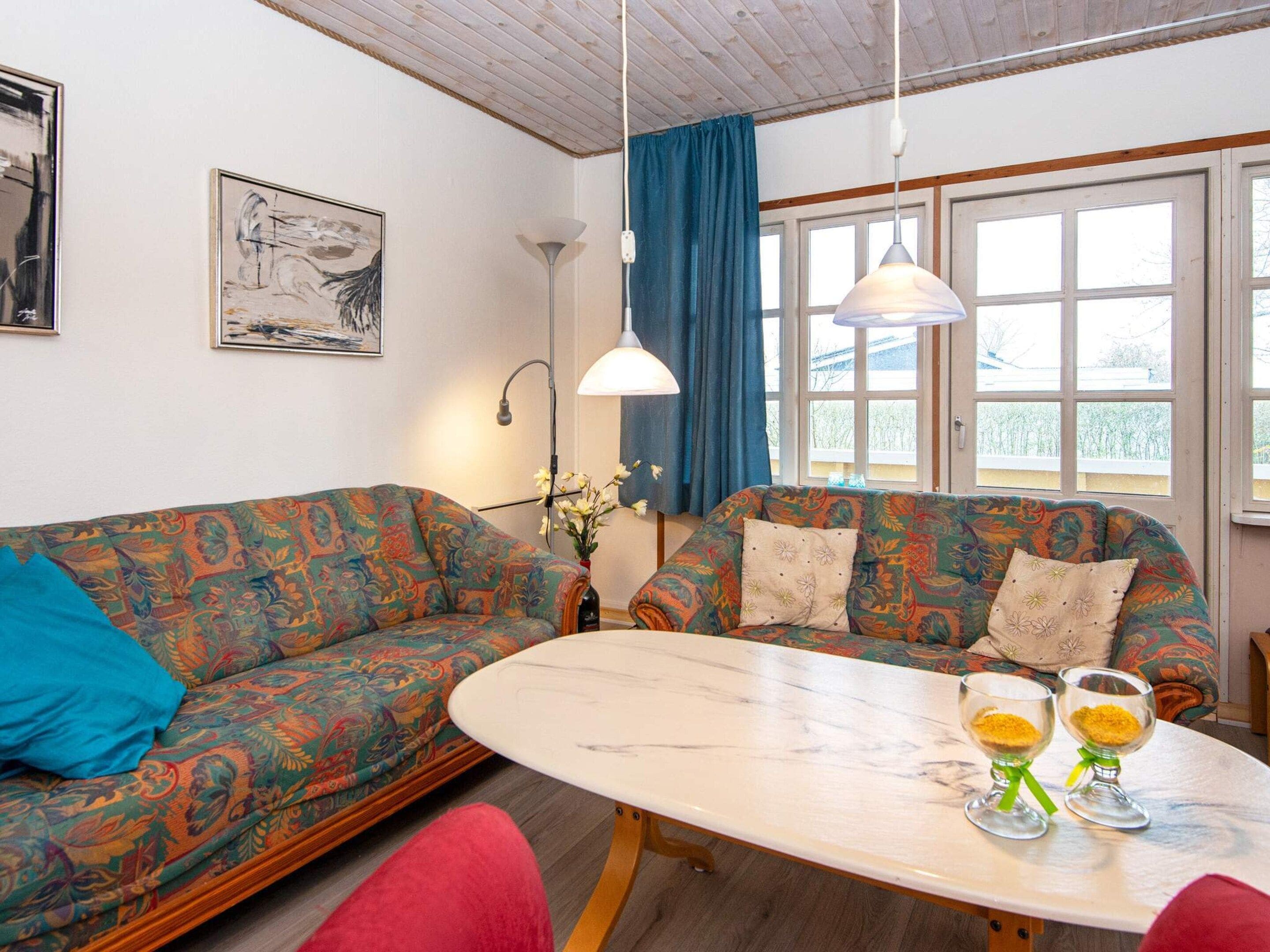 Quarto com dois sofás estampados, uma mesa redonda com dois copos e uma janela com cortinas.