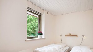 Quarto com duas camas, uma mesinha, uma janela com vista para o verde e uma estante com uma luminária.