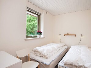Quarto com duas camas, uma mesinha, uma janela com vista para o verde e uma estante com uma luminária.