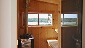 Uma cabana de madeira com sauna, uma janela com vista para uma floresta e um lago, e um pequeno banco.