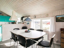 Uma sala de jantar com uma mesa branca, cadeiras pretas, uma luminária pendente e vista para o exterior através de grandes janelas.