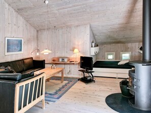 Interior acolhedor de uma cabana com teto de madeira, sofá de couro preto, mesa de jantar de madeira, televisão sobre um carrinho, lareira e uma cama com vista para o exterior.