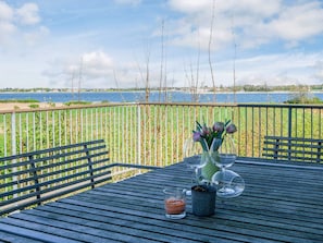 Um deck de madeira com uma mesa posta para dois, com vista para um corpo d'água e um campo gramado.