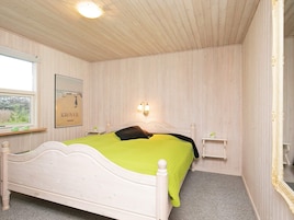 Quarto com cama branca, colcha verde, quadro na parede e mesa de cabeceira com abajur.