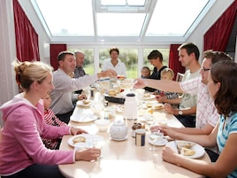 Um grupo de pessoas sentado à mesa, com comida e bebidas, em um ambiente com janelas amplas e cortinas.
