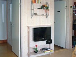 Quarto com televisão presa a parede de madeira, uma prateleira pequena com livros e uma flor, e uma porta à esquerda.