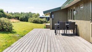 Um deck de madeira com móveis de exterior, área gramada e um edifício com janelas e porta.