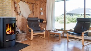 Um quarto aconchegante com lareira a lenha, duas poltronas de couro, uma mesa pequena e uma janela grande com vista para o verde.