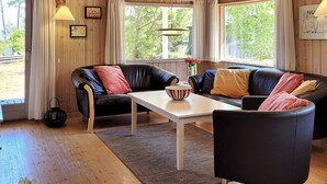 Quarto acolhedor com paredes e teto de madeira, um sofá de couro preto, uma mesa branca e janelas amplas com cortinas.