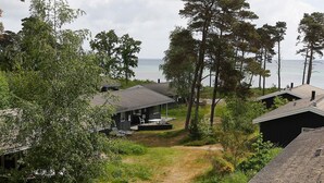 Fileira de cabanas com um caminho de terra que leva a um cais, cercada por árvores e com vista para um corpo d'água.
