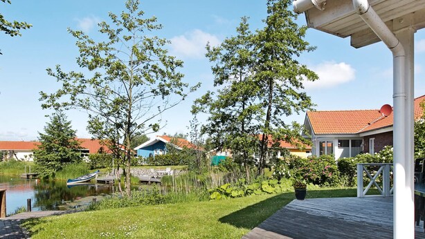 Um deck de madeira leva a uma casa com telhado de telhas vermelhas, cercada por vegetação e um pequeno lago.