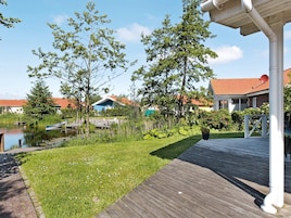Um deck de madeira leva a uma casa com telhado de telhas vermelhas, cercada por vegetação e um pequeno lago.