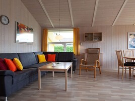 Uma sala de estar aconchegante com um sofá escuro, mesa de centro de madeira e duas cadeiras de madeira. Uma janela com cortinas permite a entrada de luz natural, e um relógio está visível na parede.