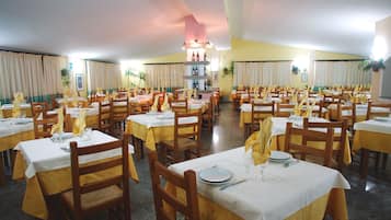 Uma ampla sala de jantar com várias mesas preparadas para servir, com cadeiras de madeira e toalhas de mesa brancas.