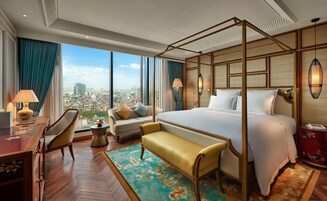 Quarto de hotel espaçoso com uma cama grande, área de estar com sofá e poltrona, escrivaninha com luminária, e vista para a cidade através de grandes janelas.