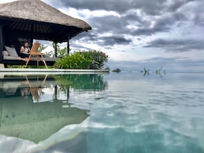 Uma piscina serena com vista infinita para o oceano, uma construção com telhado de palha e vegetação exuberante.
