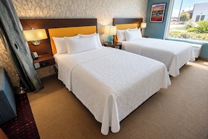 Quarto de hotel com duas camas, uma mesa de cabeceira com abajur, janela com vista e papel de parede estampado.
